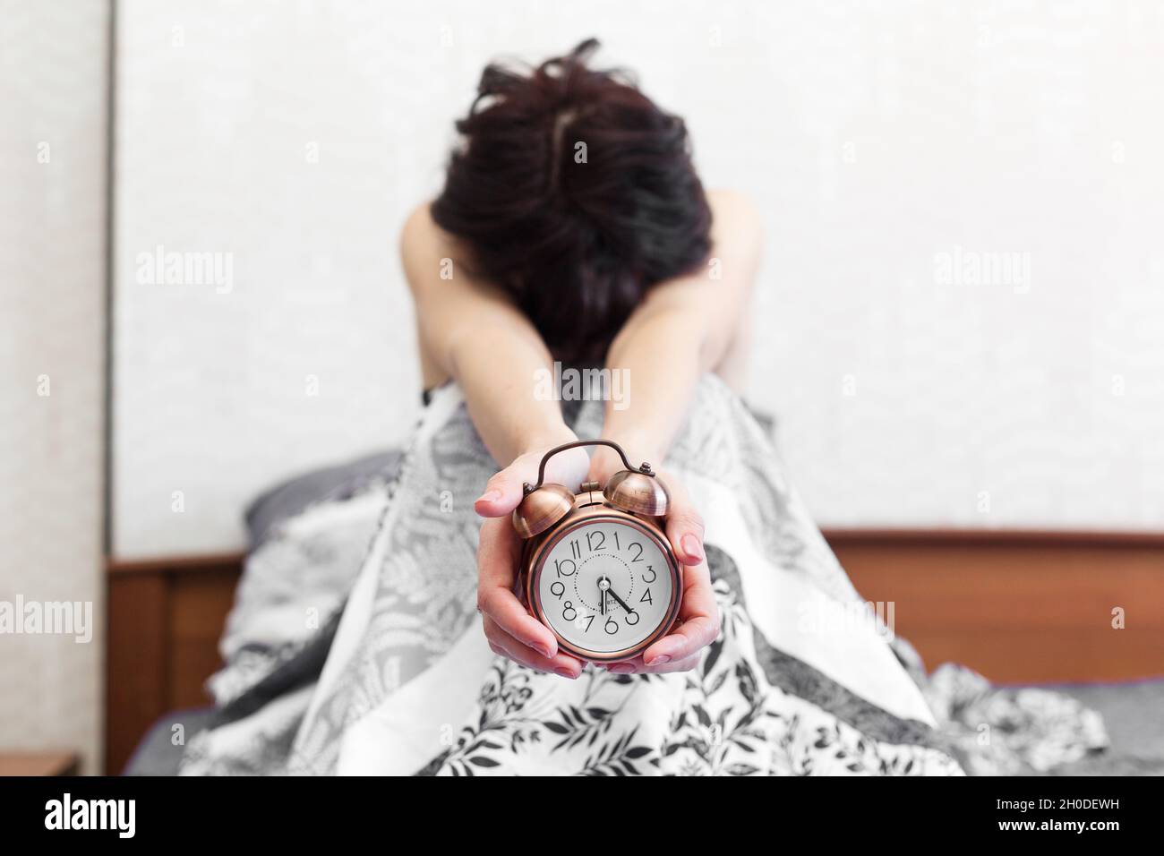 sleepy woman is sitting on the bed with an alarm clock in her hands ...
