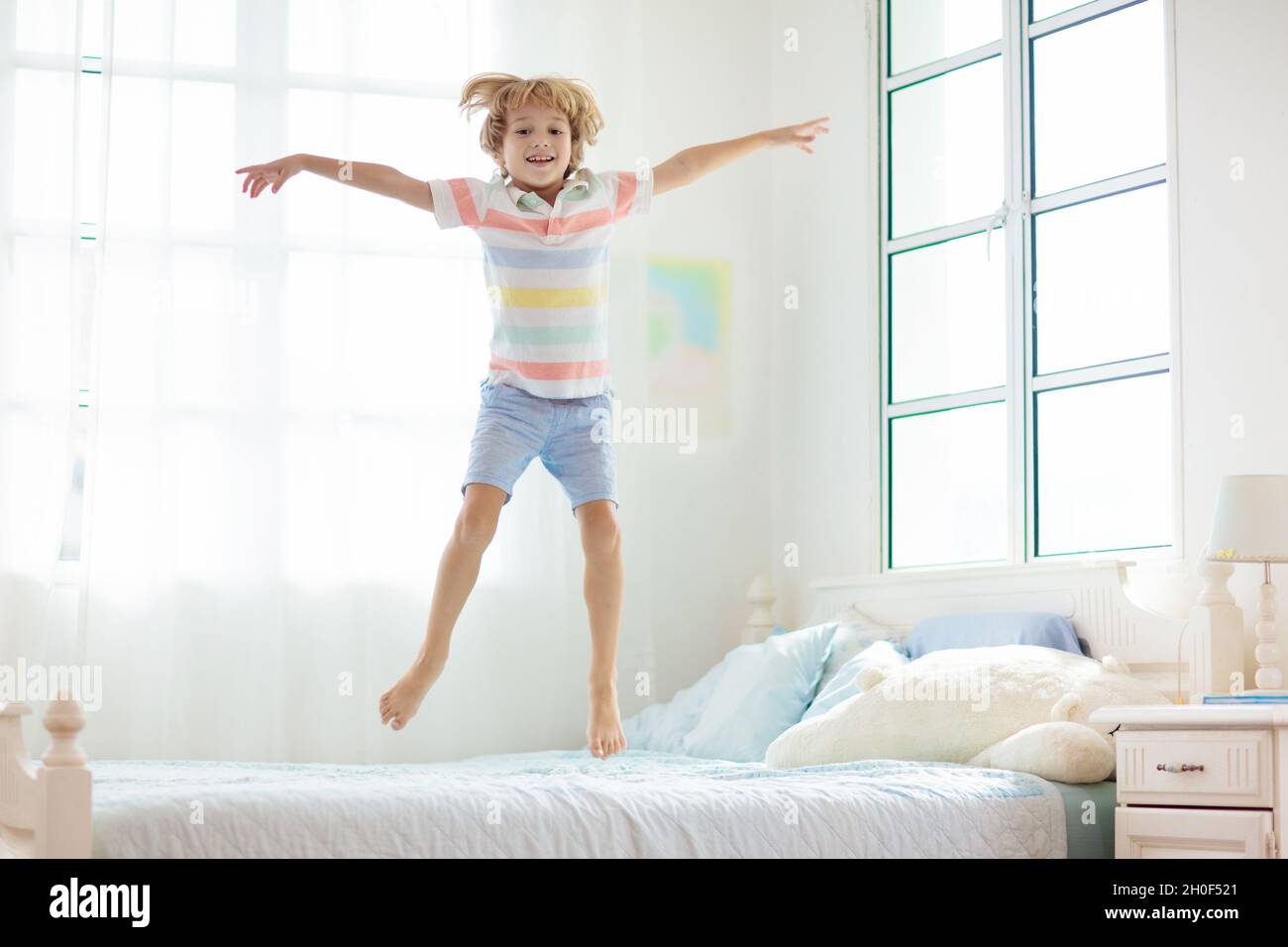 Child jumping on bed in white bedroom with big window. Kids play at ...