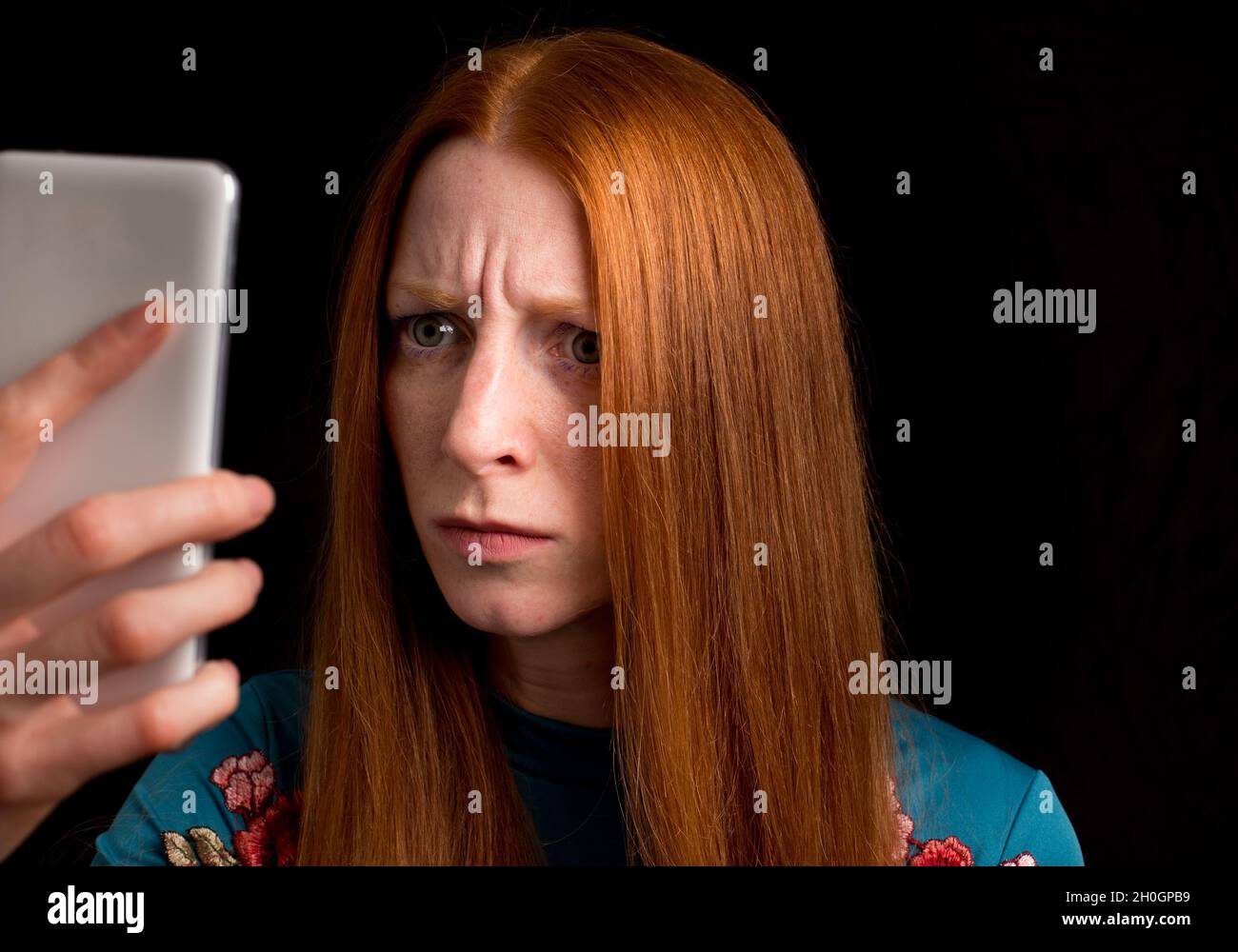 Angry redhead girl looking at mobile phone against black background ...