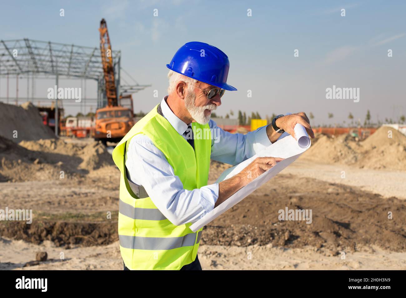 Senior engineer looking at blueprints in front of metal construction at ...
