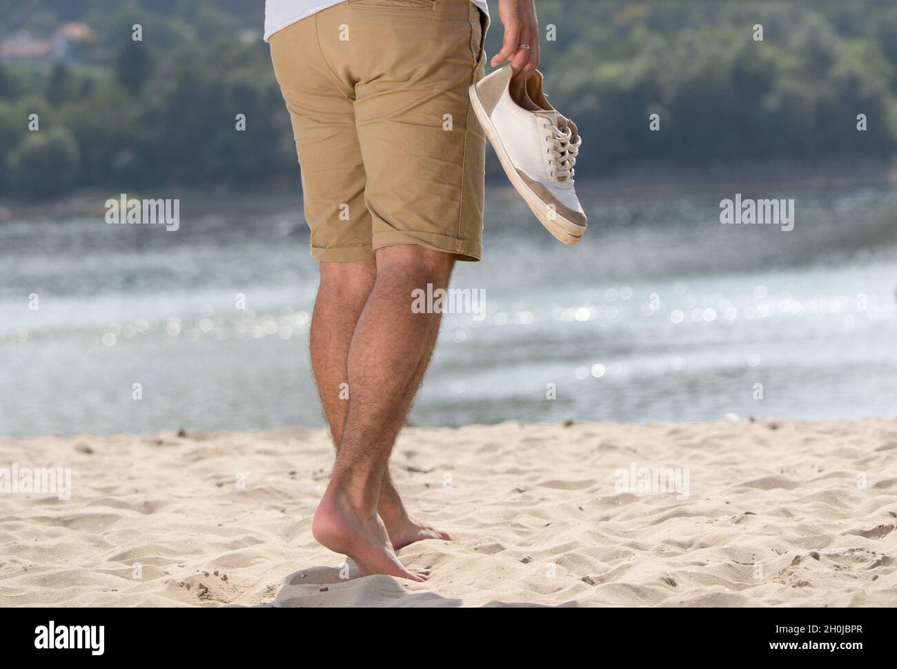 Man walking barefoot with shoes in hand on sand at river coast ...