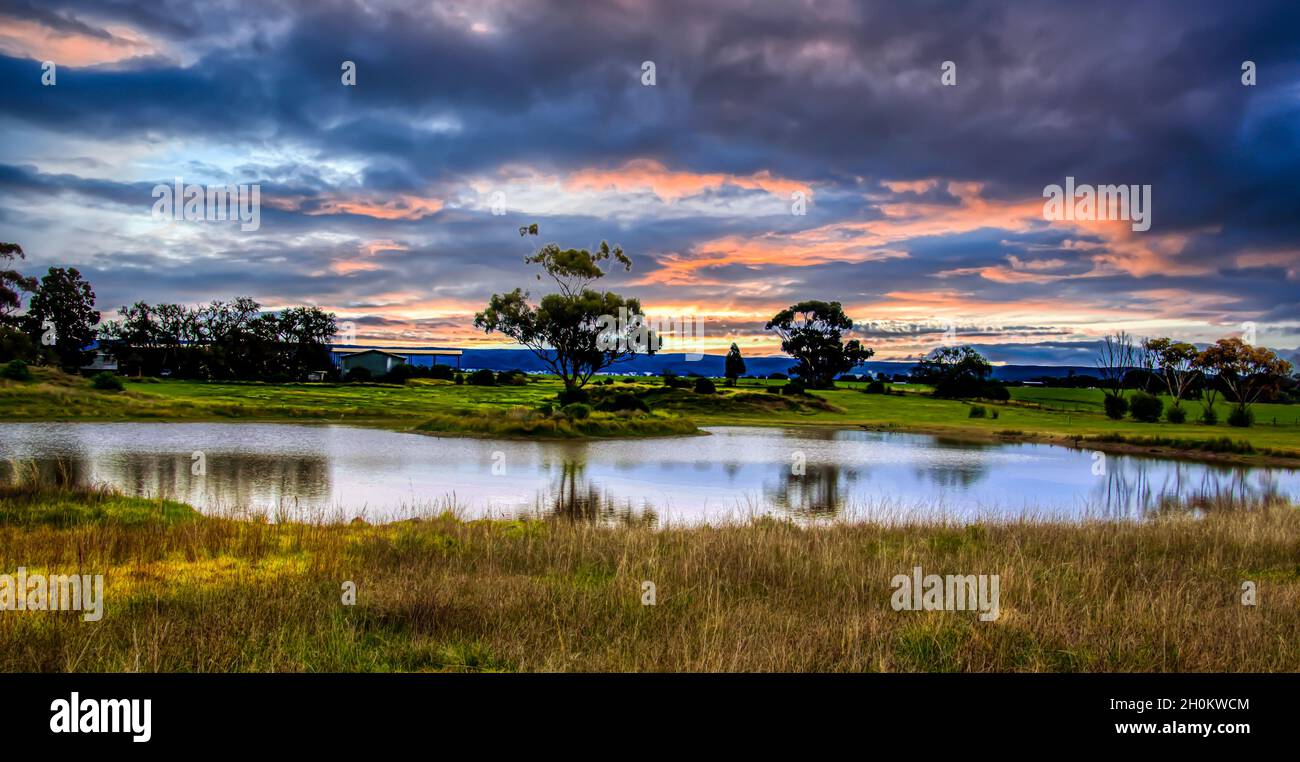 Shot of a lake near a grassy field with trees under a cloudy sunset ...