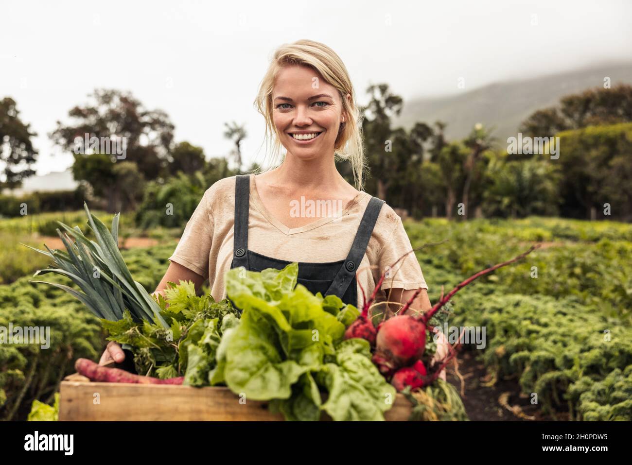 Vegetable farmer smiling cheerfully after harvest. Happy young woman holding a box full of ...