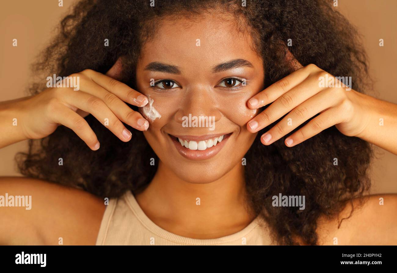 Smiling dark skinned woman with curly afro hair applies face cream on ...