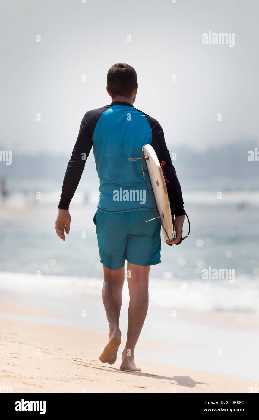 Rear view of handsome man carrying surfboard on sand beach to water ...