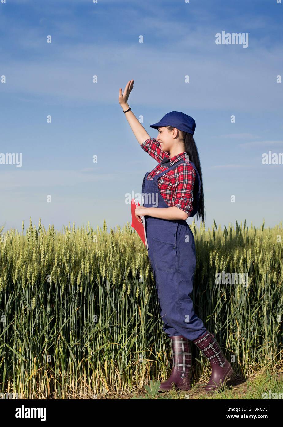 Pretty young farmer woman standing in front of green wheat field and ...