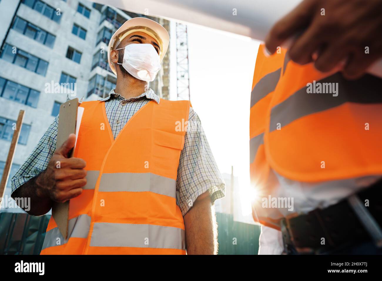 Two men in workwear and medical masks working with blueprints on object ...