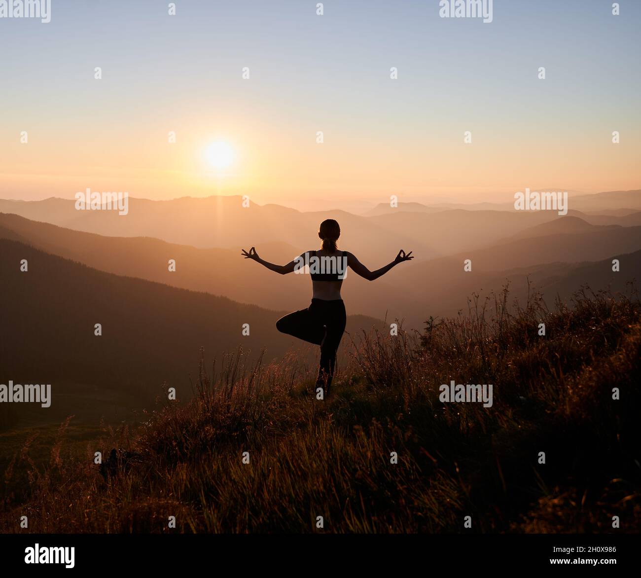 Back view of slim woman performing yoga pose on grassy hill with orange ...