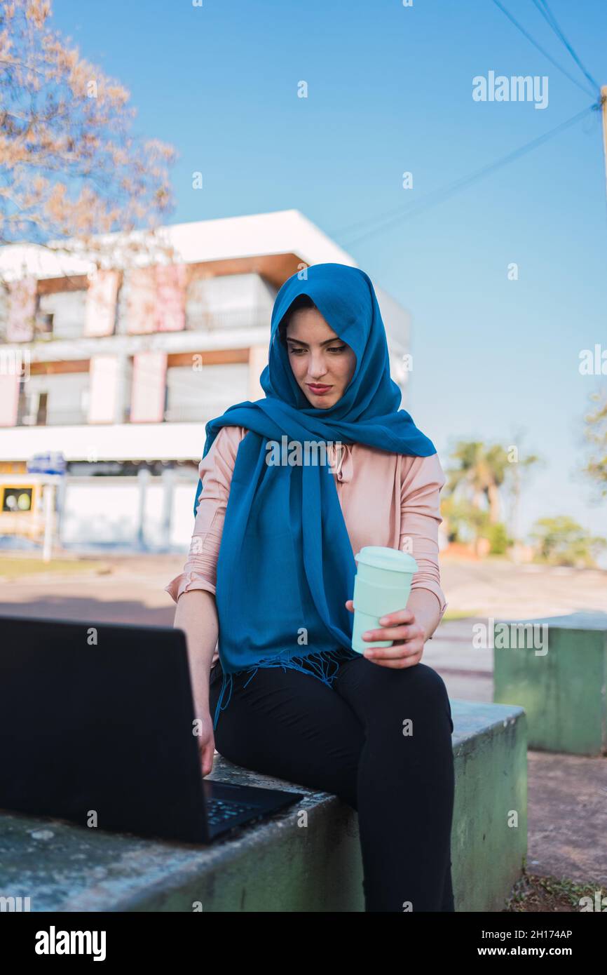Smiling Arab female freelancer in hijab sitting on bench with takeaway ...