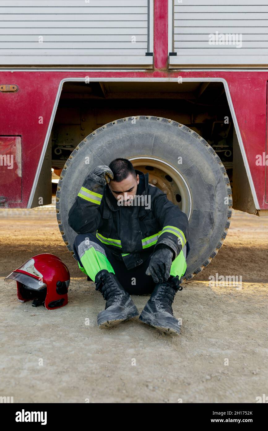 Exhausted male firefighter wearing uniform sitting on ground near fire ...