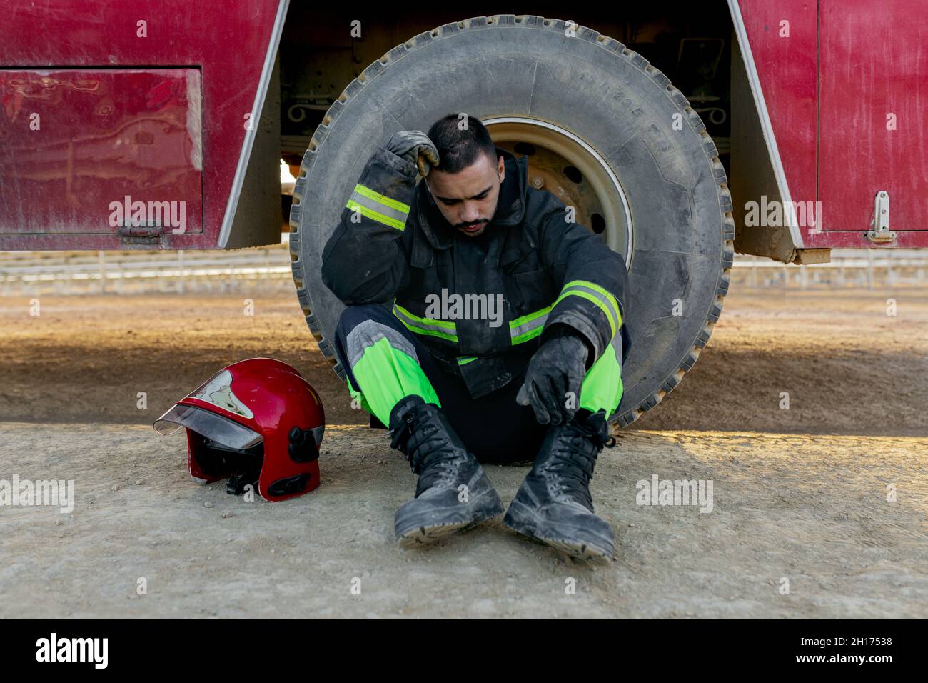 Exhausted male firefighter wearing uniform sitting on ground near fire ...