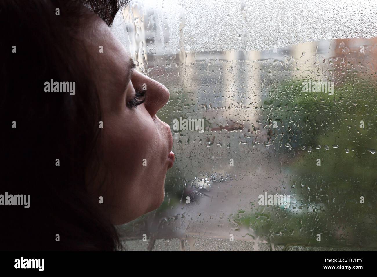 A sad woman blows air with her lips on a window fogged from the rain ...