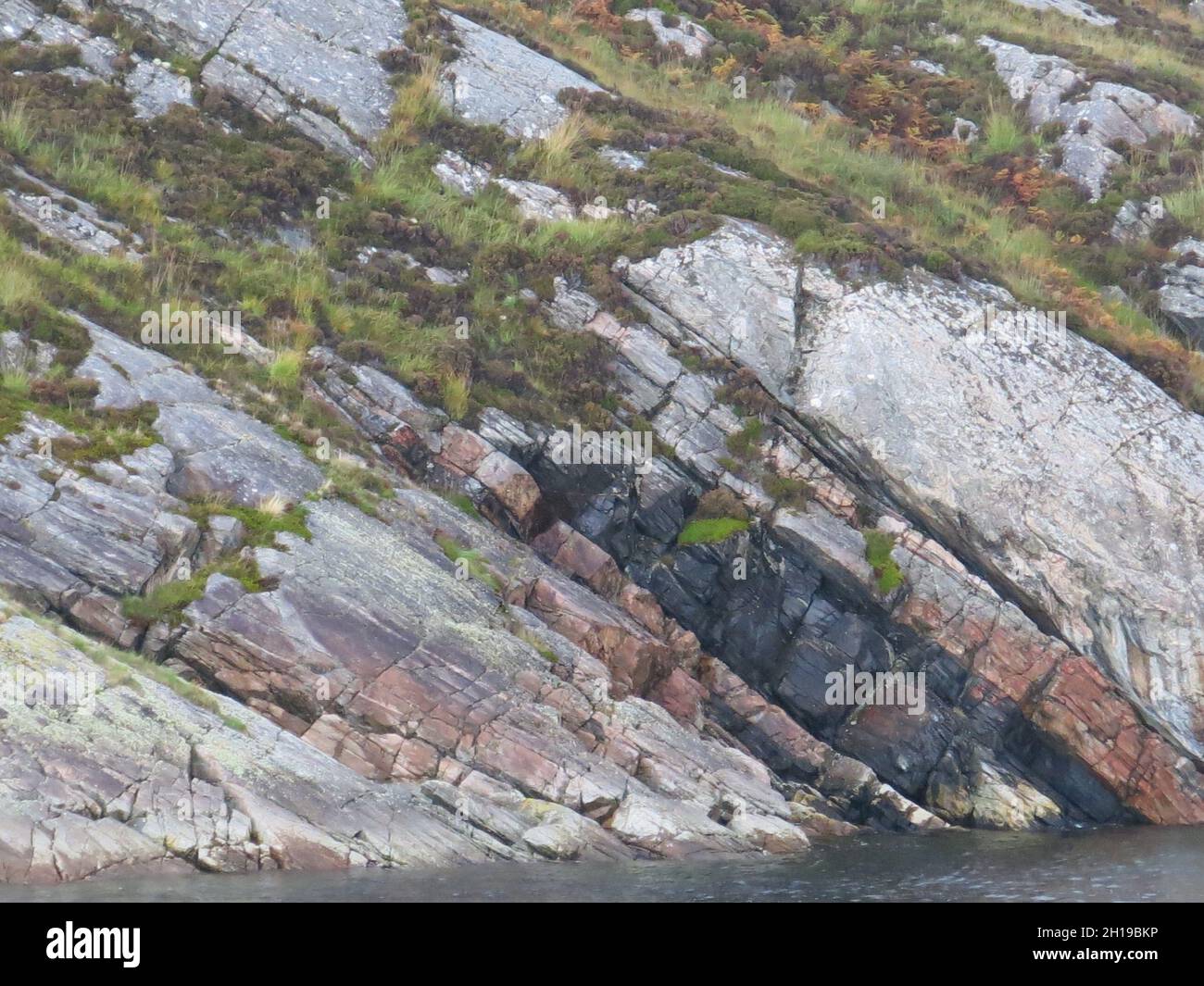 Rocky outcrop with grasses and vegetation sloping down to the water's ...