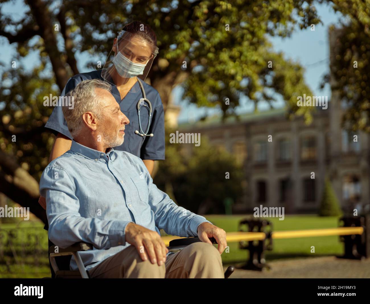 Caring young nurse wearing face shield and mask talking to mature man ...