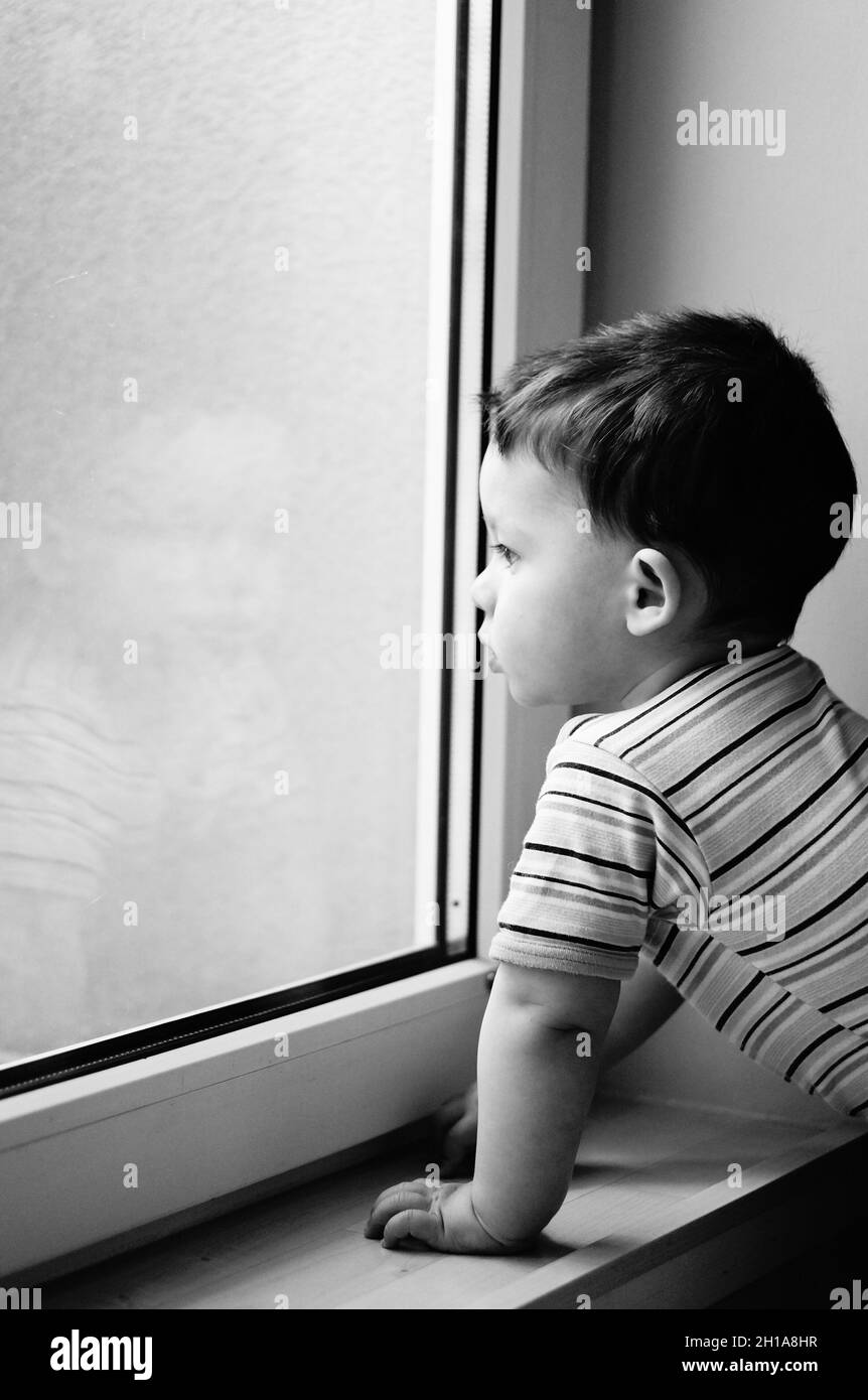 Grayscale of a Caucasian male kid looking outside through glass window ...