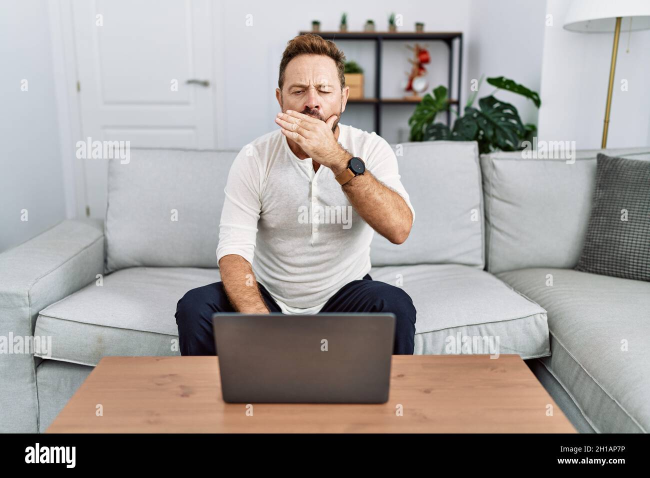 Middle age man using laptop at home bored yawning tired covering mouth ...