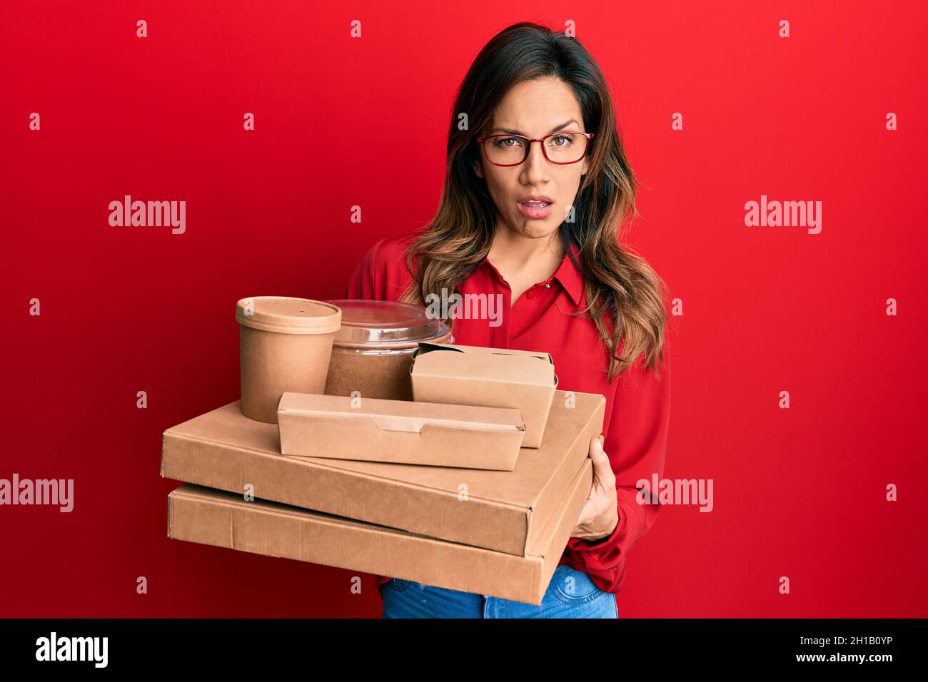Young latin woman holding take away food in shock face, looking ...