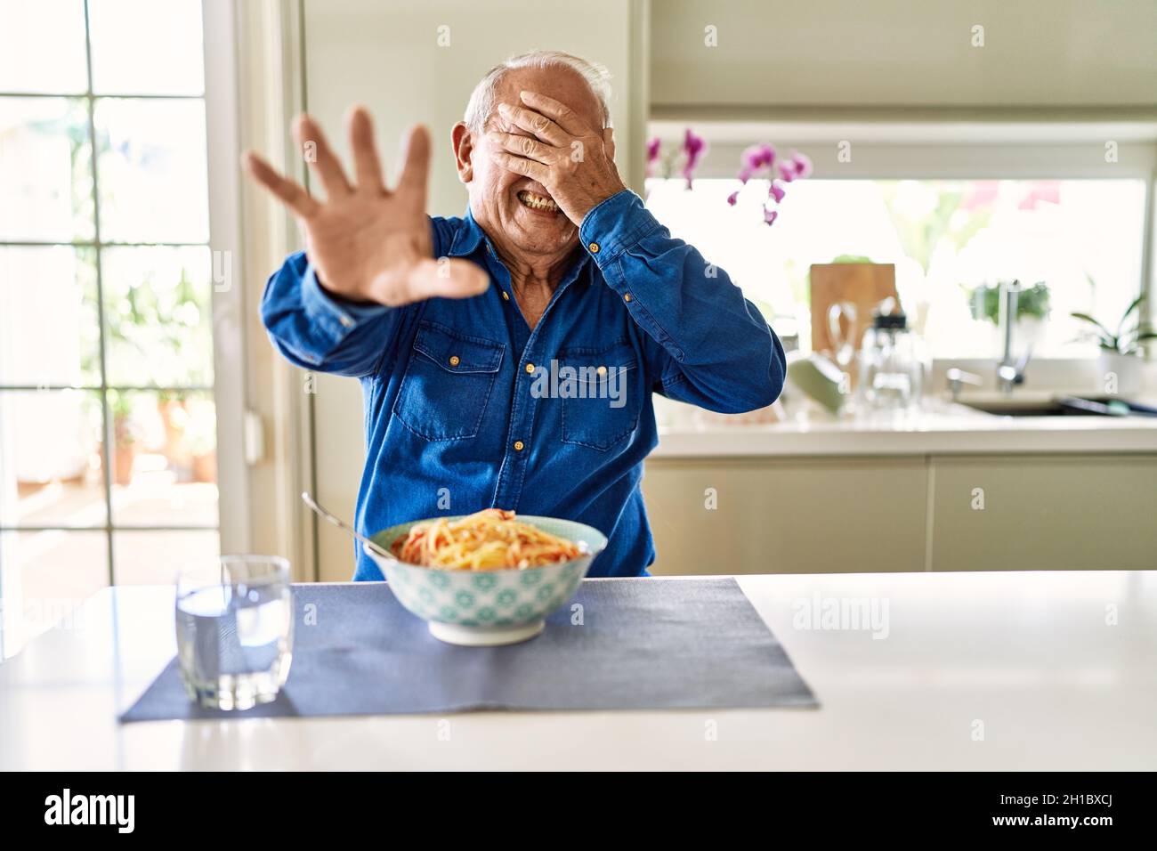 Senior man with grey hair eating pasta spaghetti at home covering eyes ...