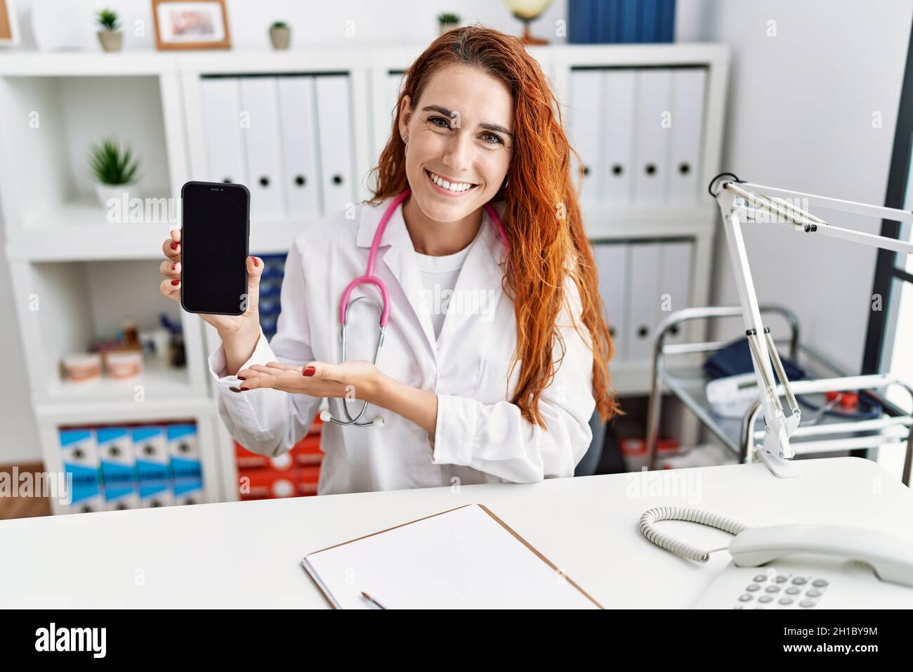 Young redhead doctor woman holding smartphone showing blank screen ...