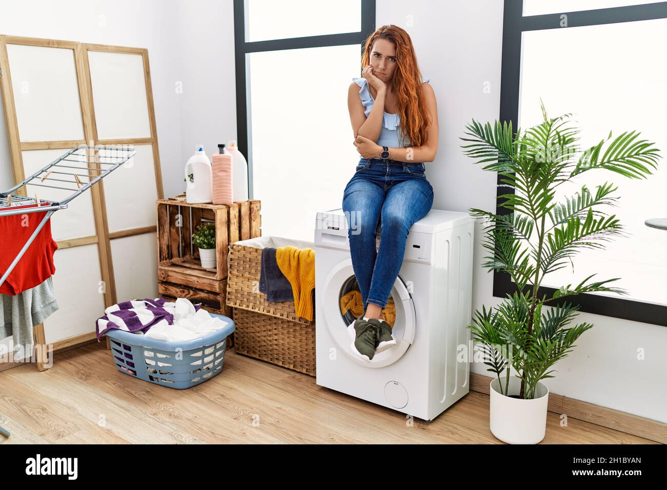 Young redhead woman doing laundry sitting on washing machine thinking ...