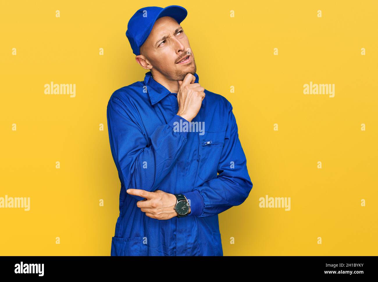 Bald man with beard wearing builder jumpsuit uniform thinking worried ...