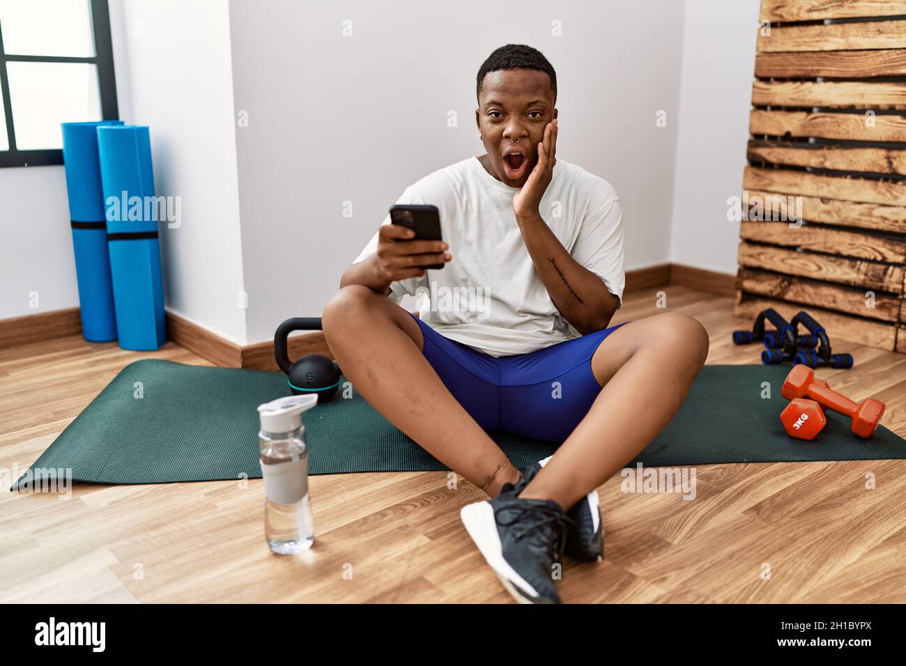 Young african man sitting on training mat at the gym using smartphone ...