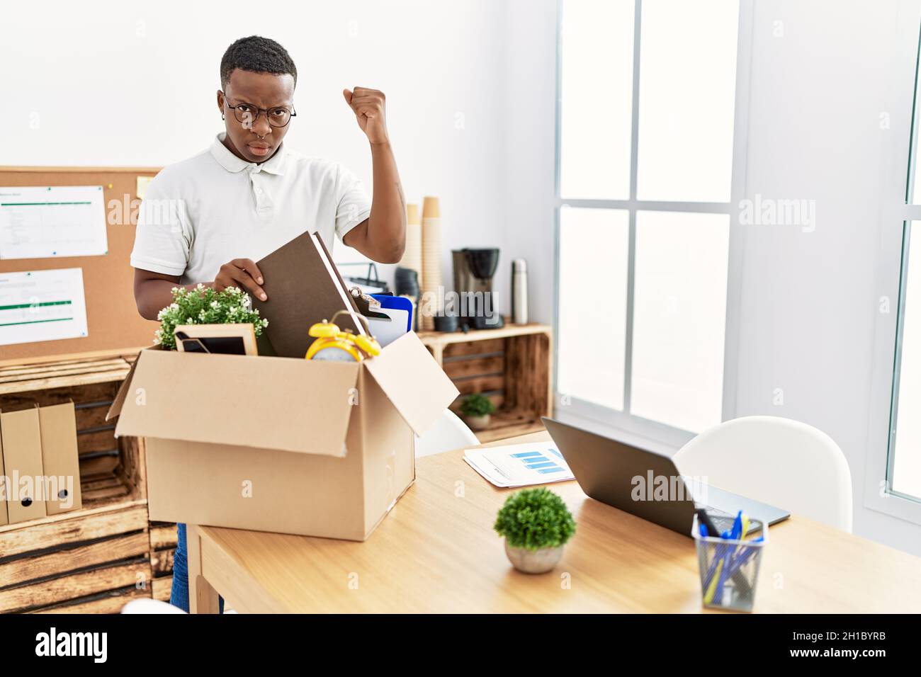 Young african man putting office objects into cardboard box annoyed and ...