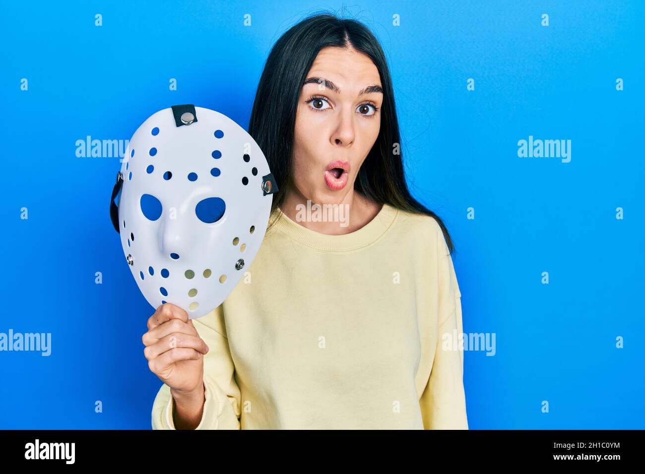 Young brunette woman holding hockey mask scared and amazed with open ...