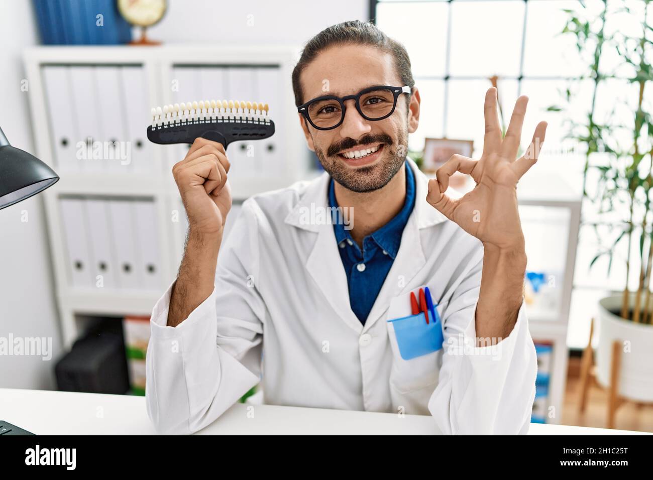 Young hispanic dentist man comparing teeth whitening doing ok sign with ...
