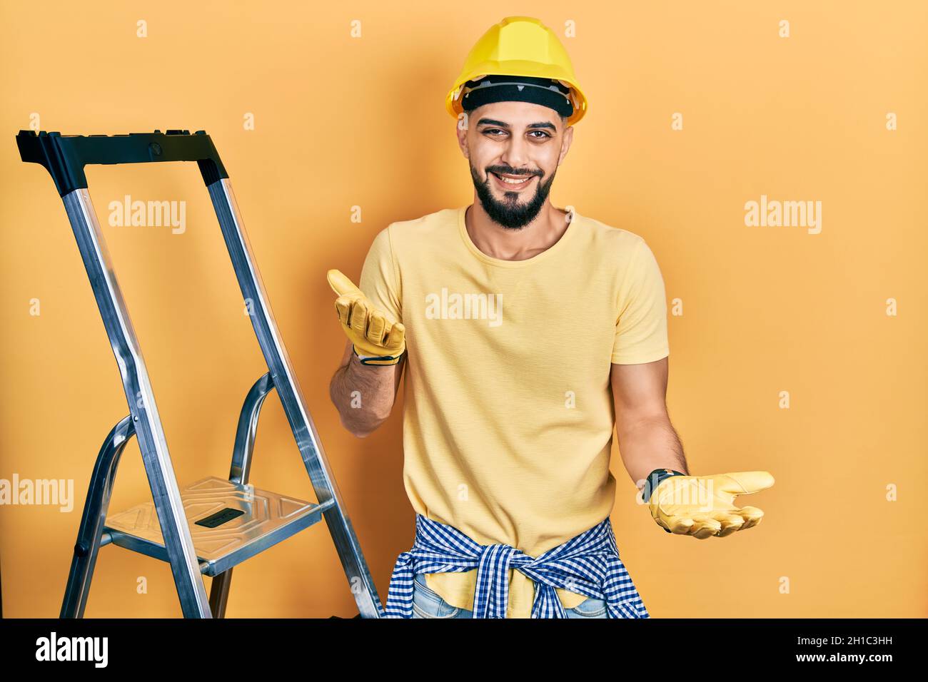 Handsome man with beard by construction stairs wearing hardhat smiling cheerful with open arms ...