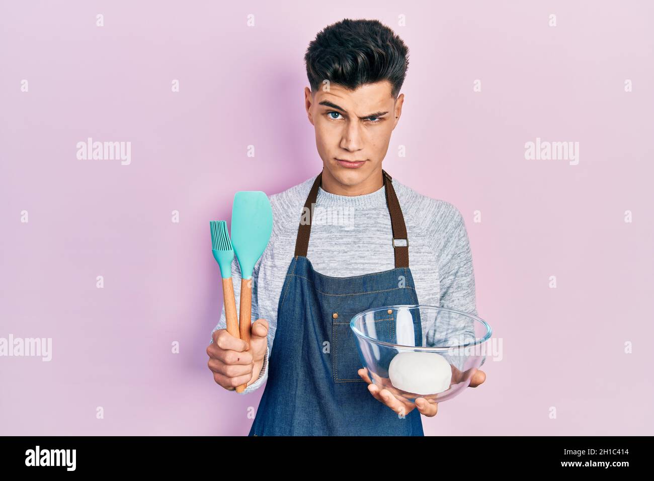 Young hispanic man holding bread dough and cooking tools skeptic and ...