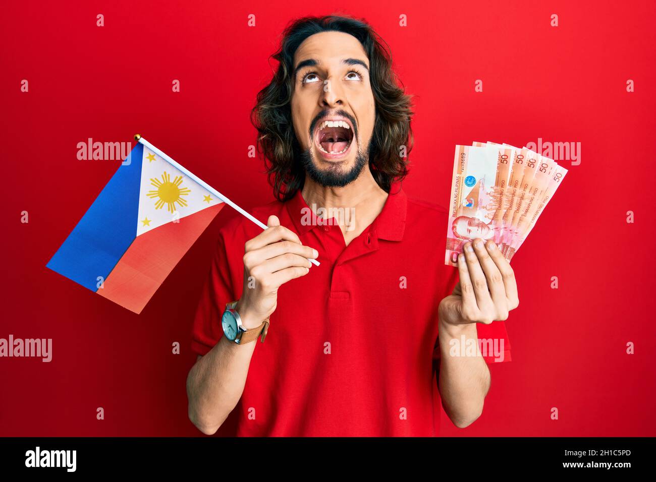 Young hispanic man holding philippines flag and pesos banknotes angry ...