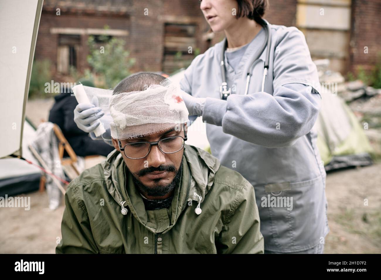 Young doctor in uniform putting bandage around wounded head of refugee ...