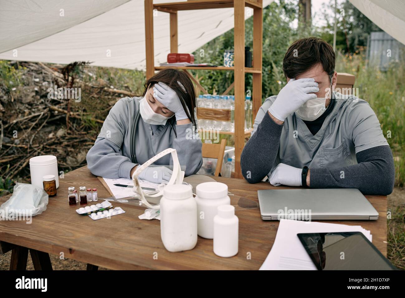 Two young exhausted clinicians in protective workwear sitting by table ...