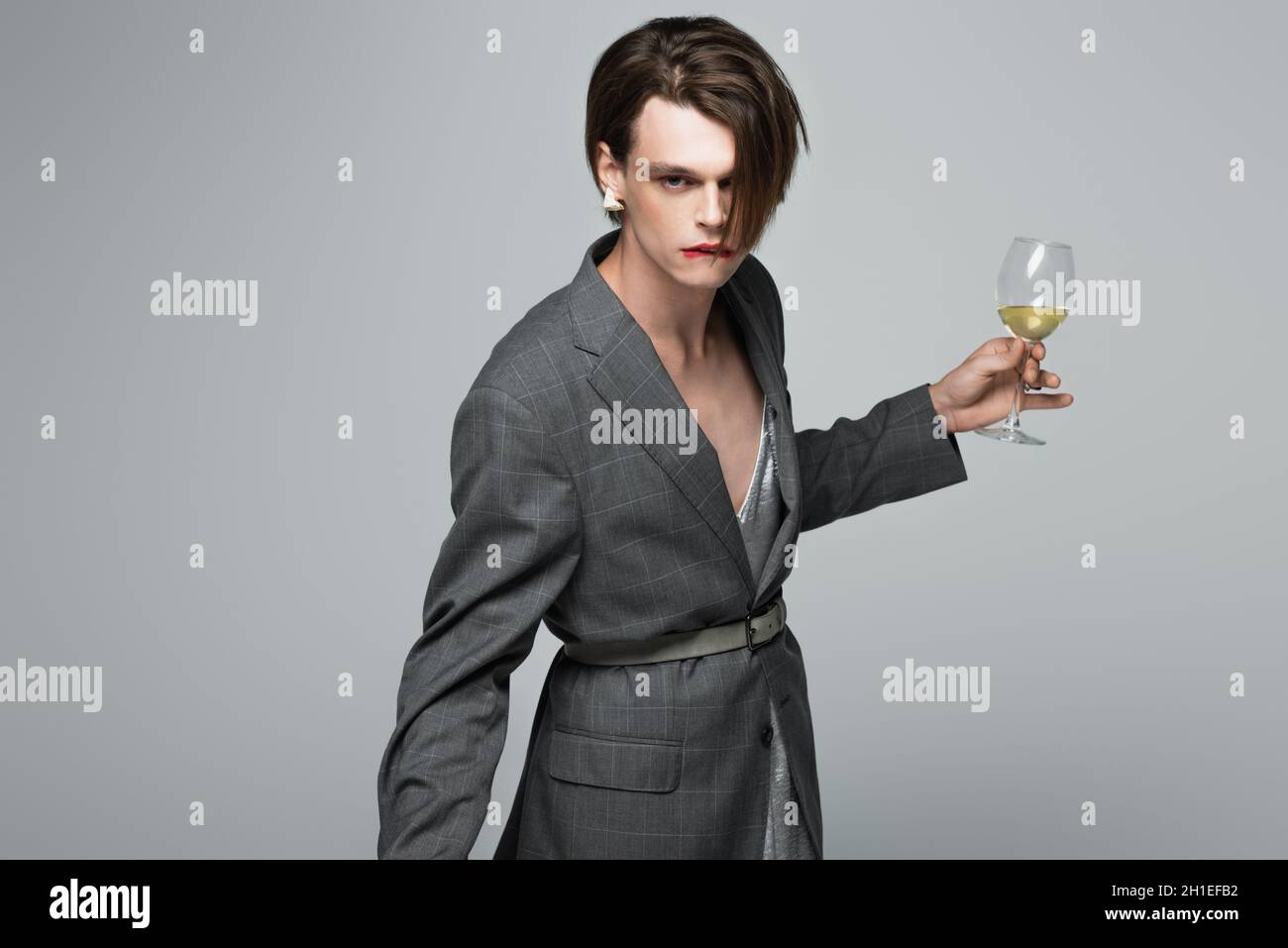 young transgender man in blazer biting lip while holding glass of wine ...