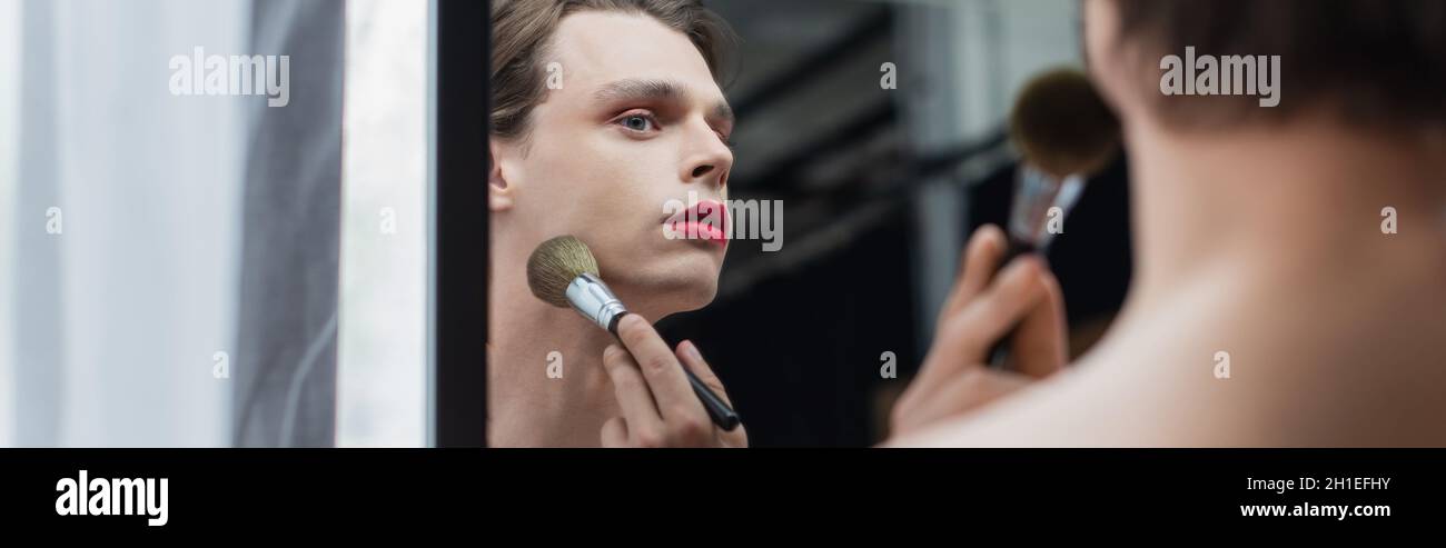 young transgender man applying face powder on neck near mirror, banner ...