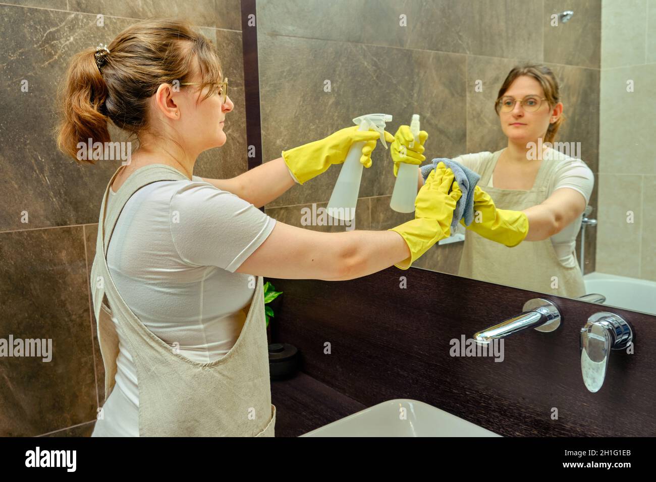 A woman is cleaning the bathroom by wiping the mirror with a rag ...