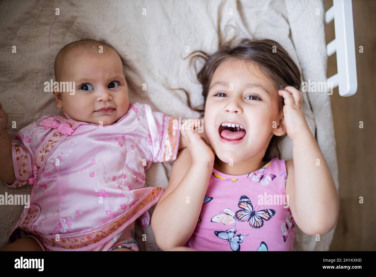 Confused baby girl wearing pink Asian attire lying down next to laughing older sister. Two ...