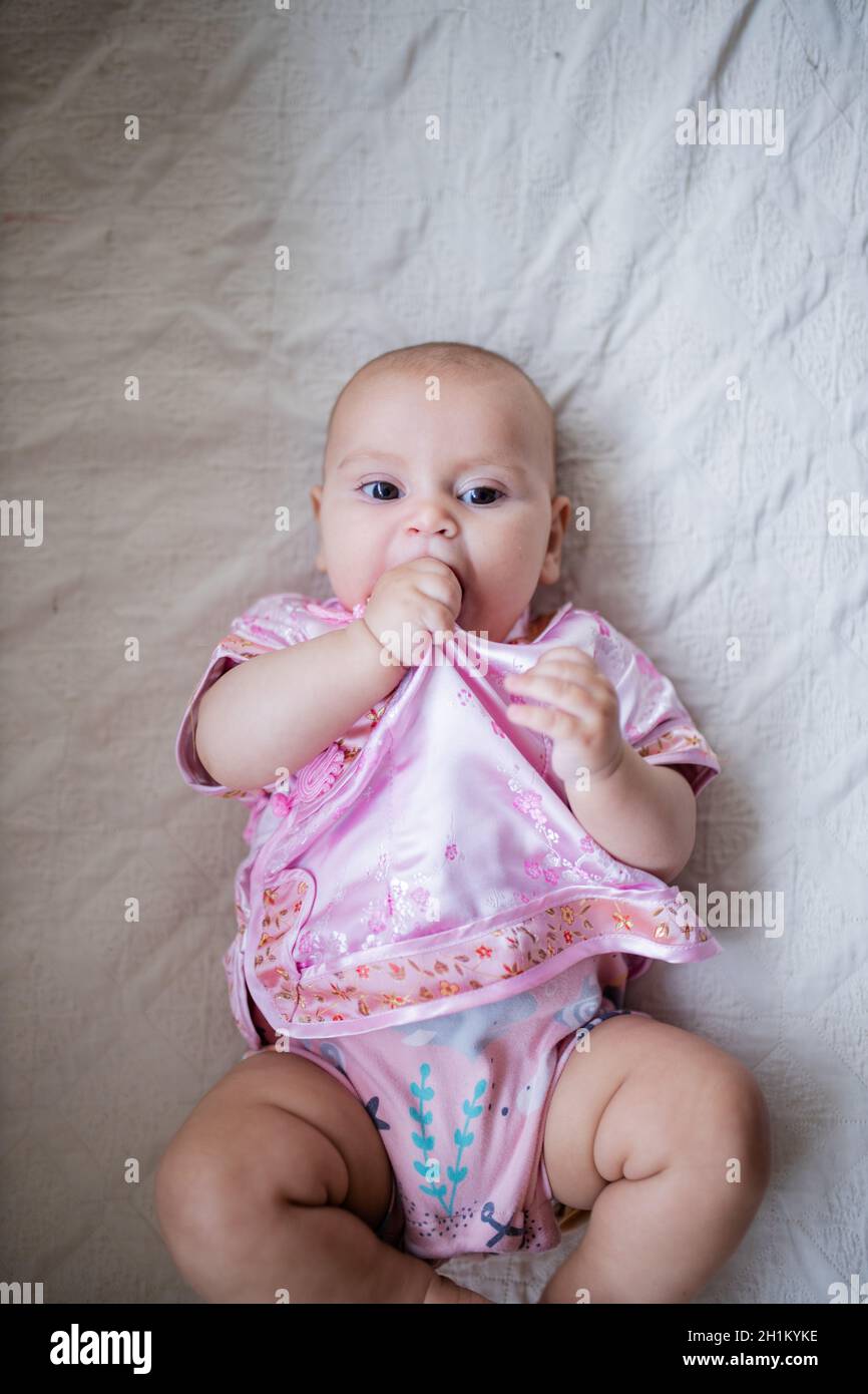 Adorable baby girl from above biting her Asian pink attire and lying ...