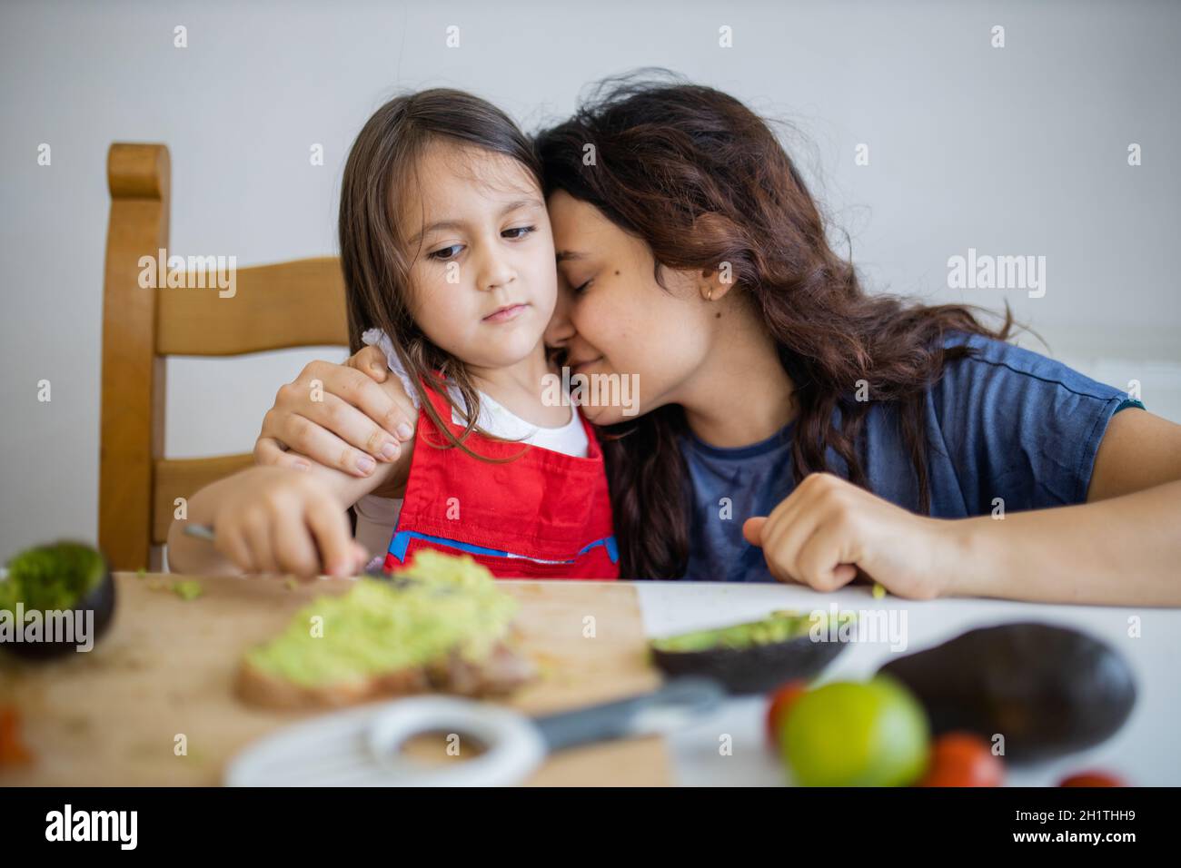 Happy mother lovingly hugging daughter while spreading avocado on toast ...