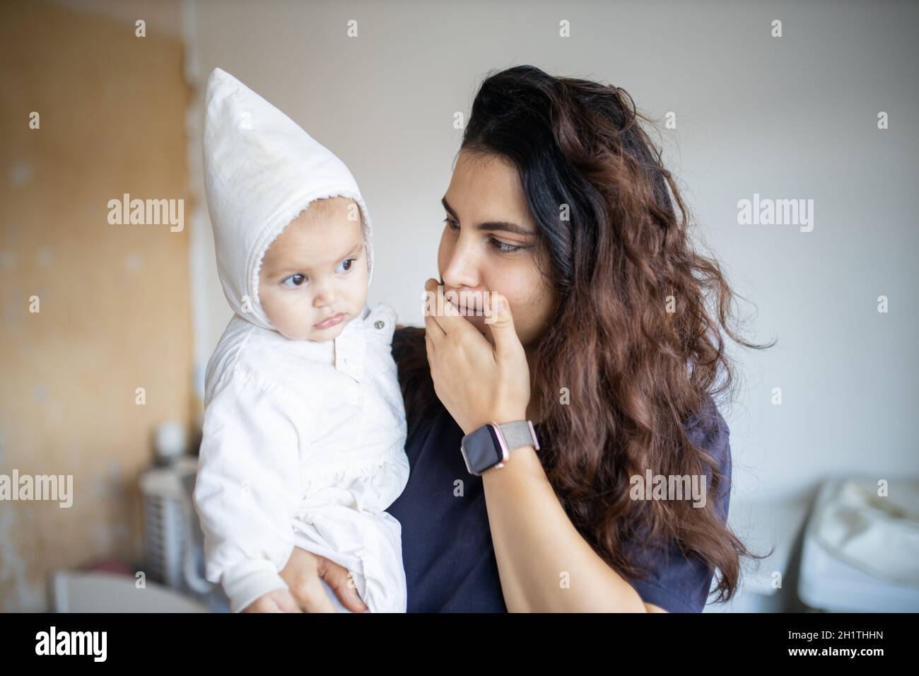 Concerned brunette woman holding her adorable baby in white clothing ...