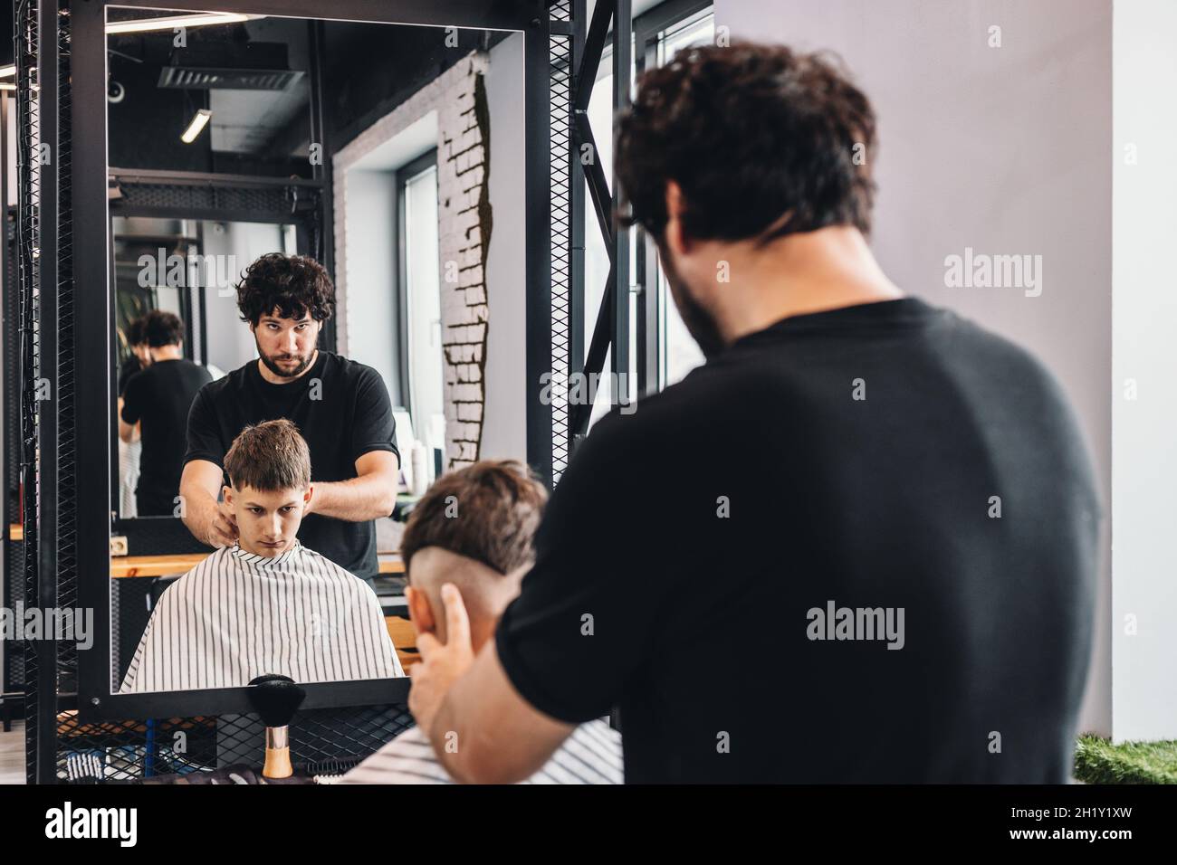 Handsome tall bearded brunette barber gives a trendy haircut to a ...