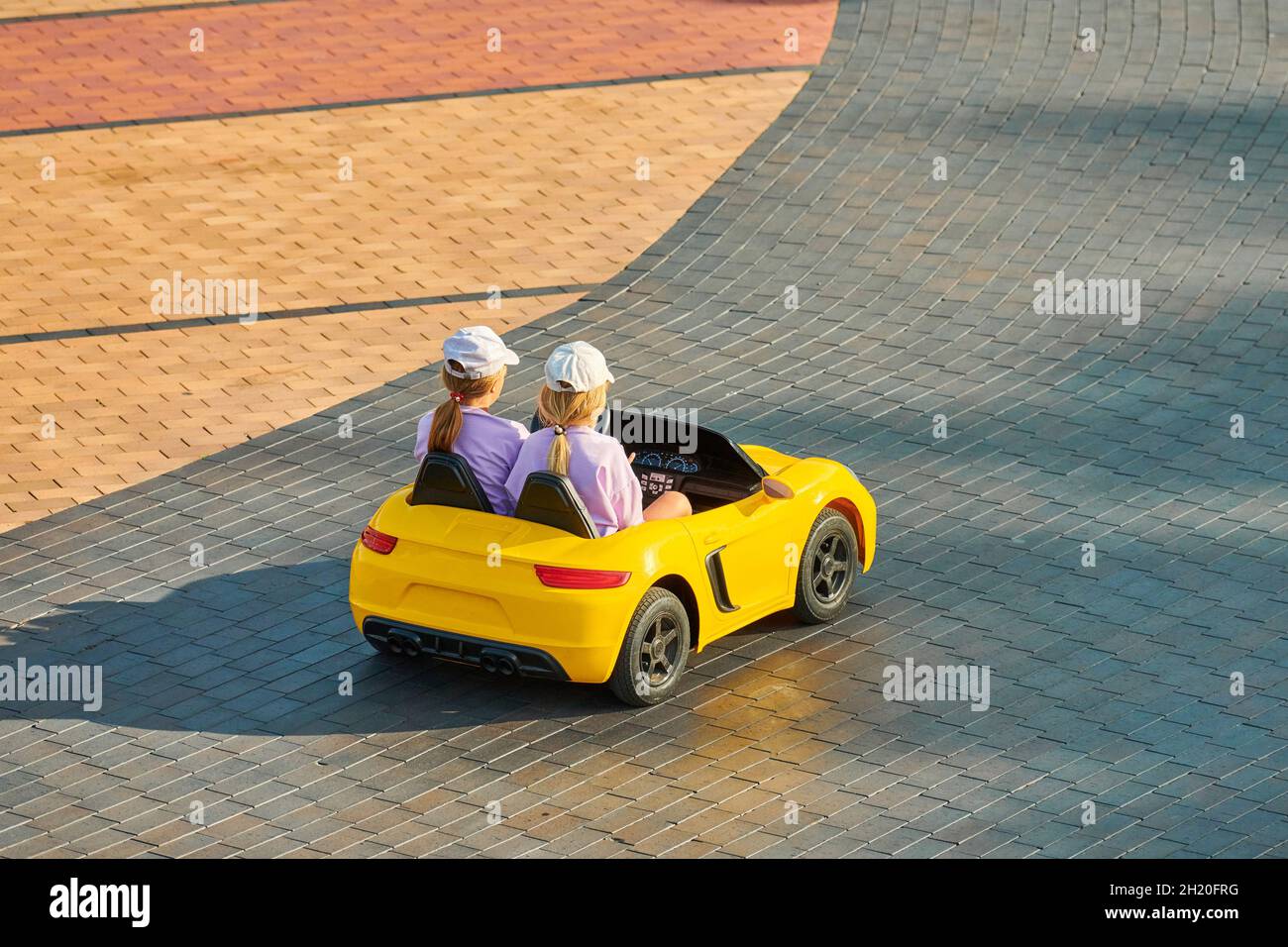 Two little girls driving yellow toy car on pavement in amusement park ...
