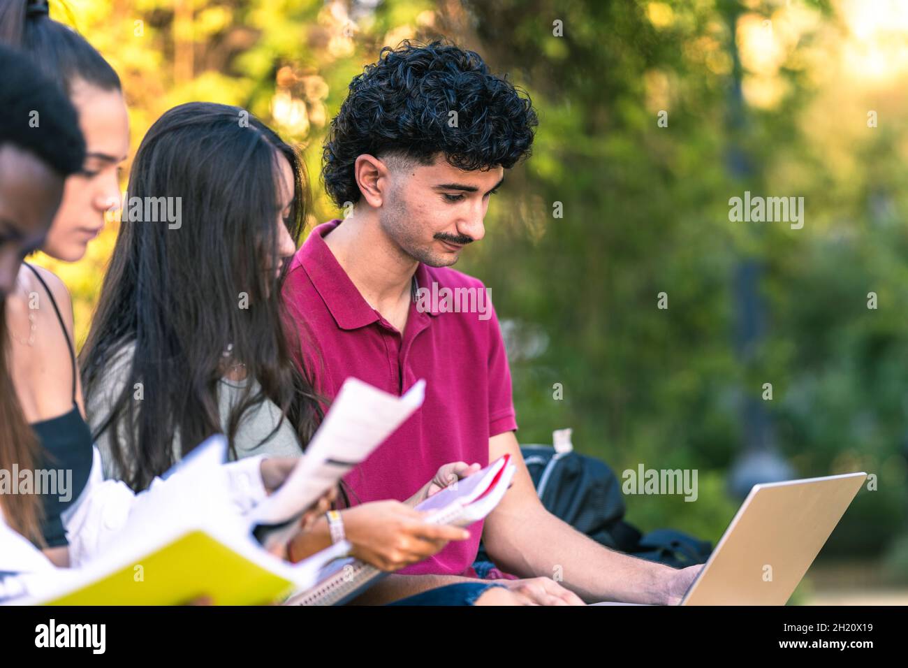 Diverse young people studying with laptop and papers sitting on a bench ...