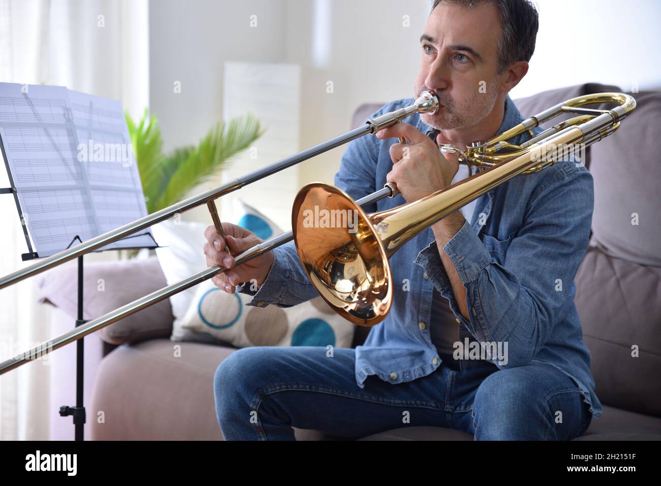 Man practicing the trombone at home sitting on the sofa. Front view ...