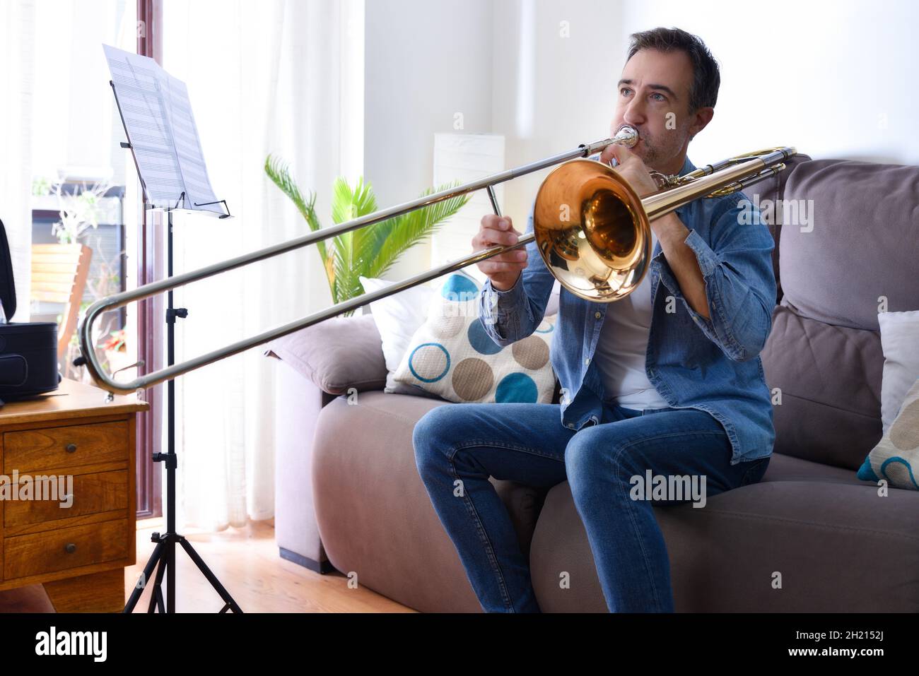Man practicing the trombone at home sitting on the sofa. Front view ...