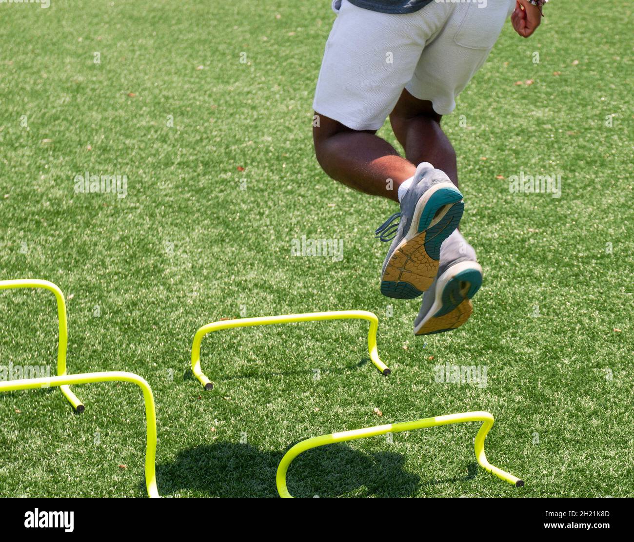 A high school track runner jumping on one leg over yellow mini hurdles on a green turf field ...
