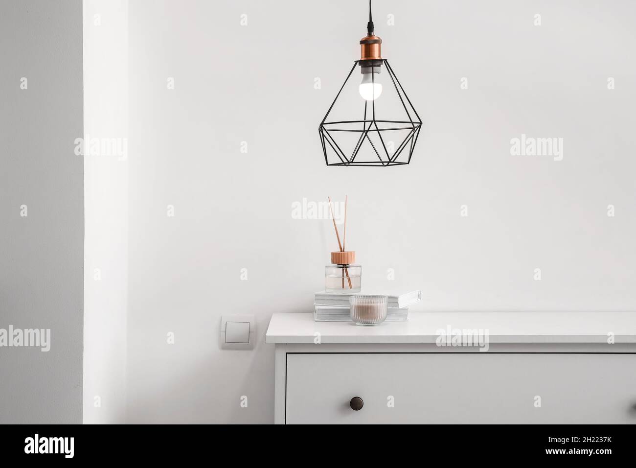Reed diffuser with books on shelf and glowing lamp near white wall ...