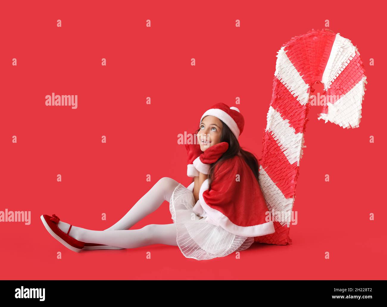 Little girl in Santa hat and with candy cane pinata on color background ...