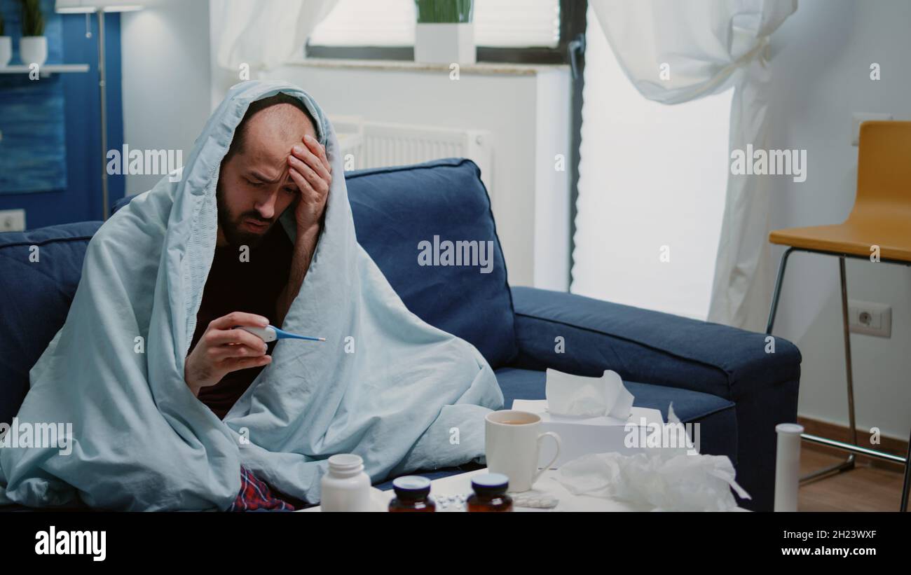 Ill person wrapped in blanket looking at thermometer for temperature ...